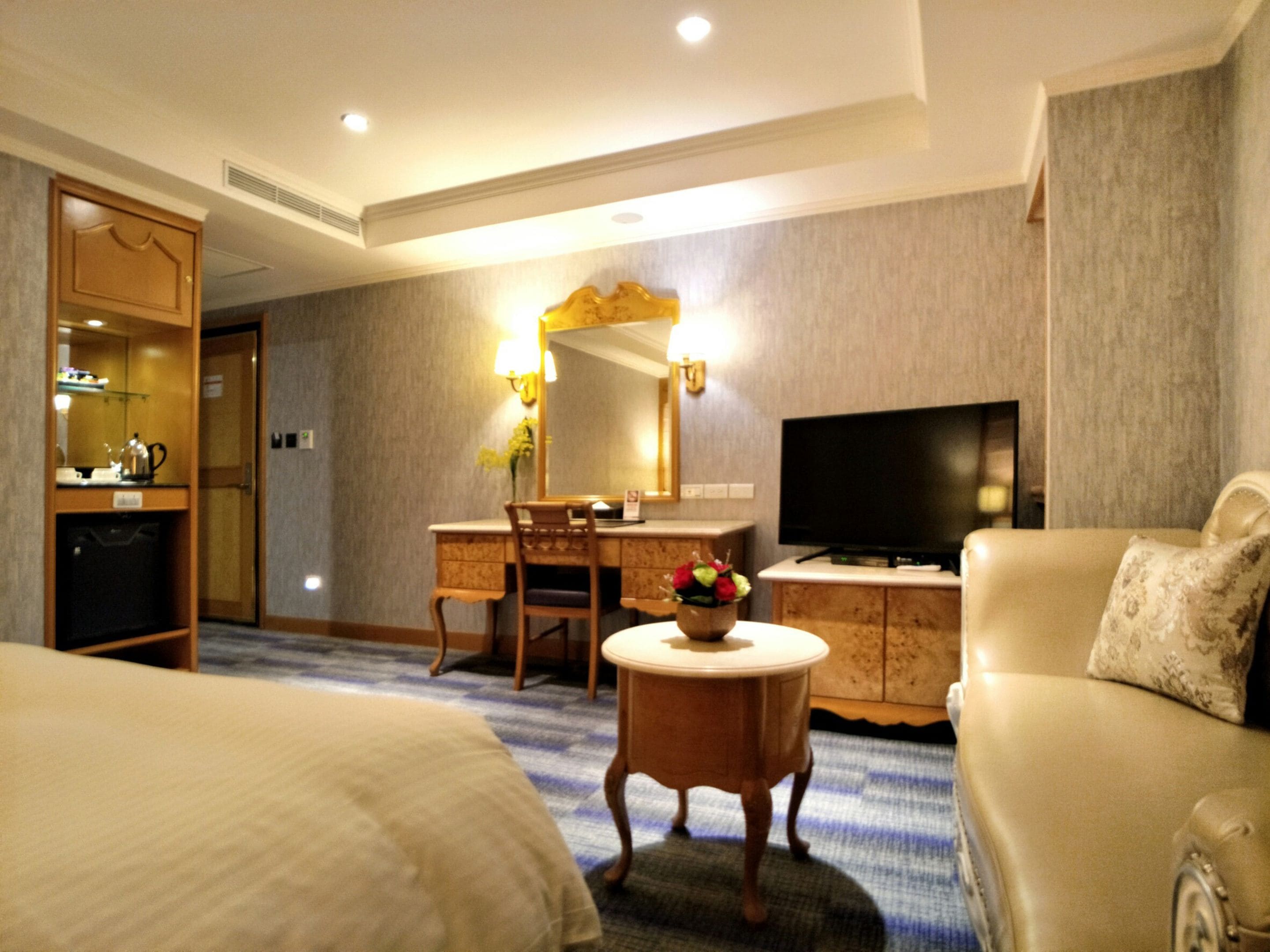 Quarto de hotel com cama, sofá, escrivaninha com cadeira, televisão e espelho.