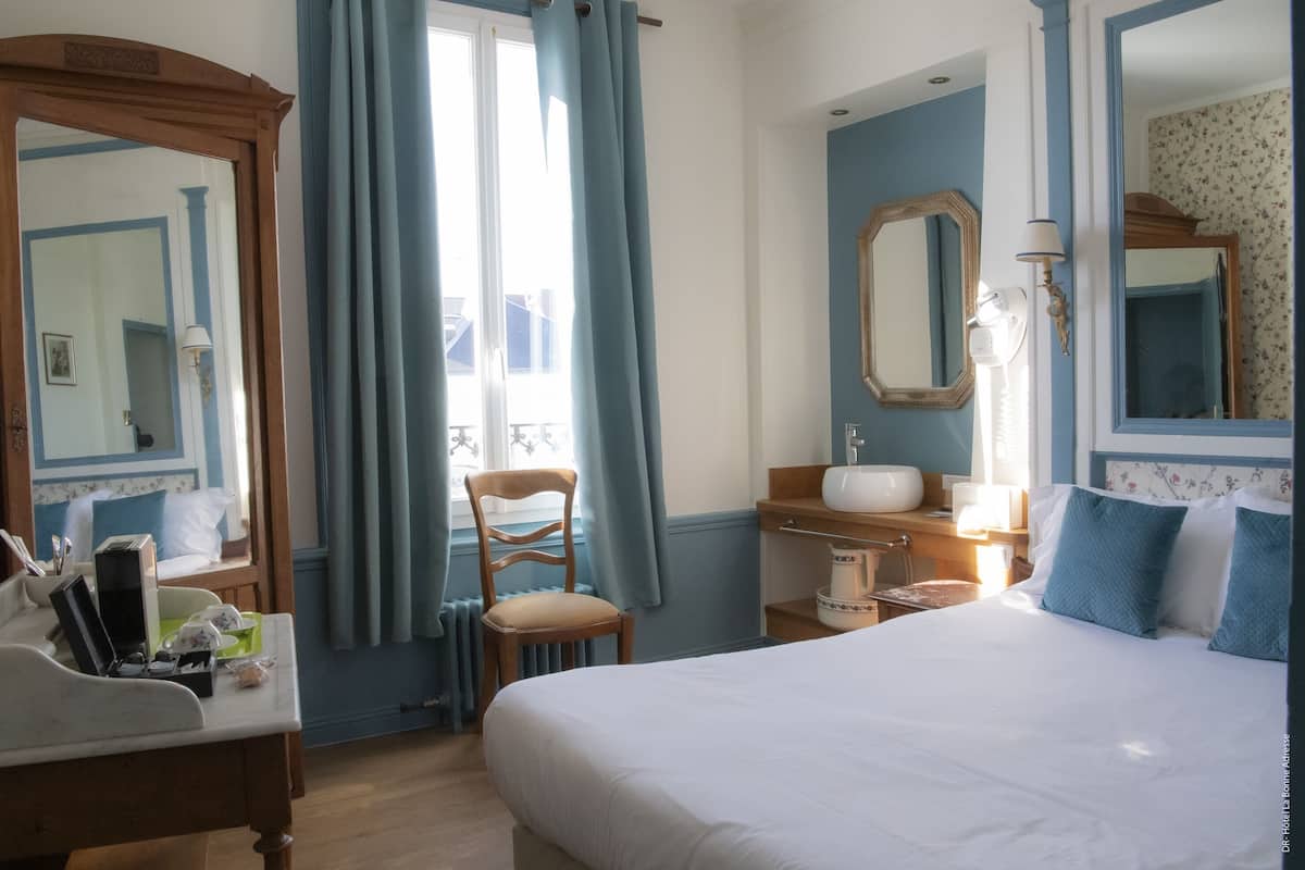 Quarto de hotel com cama, cadeira, escrivaninha com luminária, espelho, janela com cortinas e banheiro com pia e espelho.