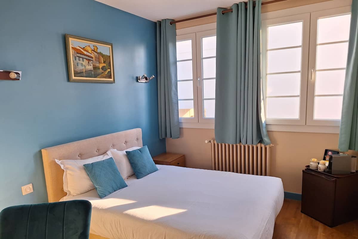 Quarto com cama, cadeira, mesa de cabeceira, radiador e janela com cortinas.
