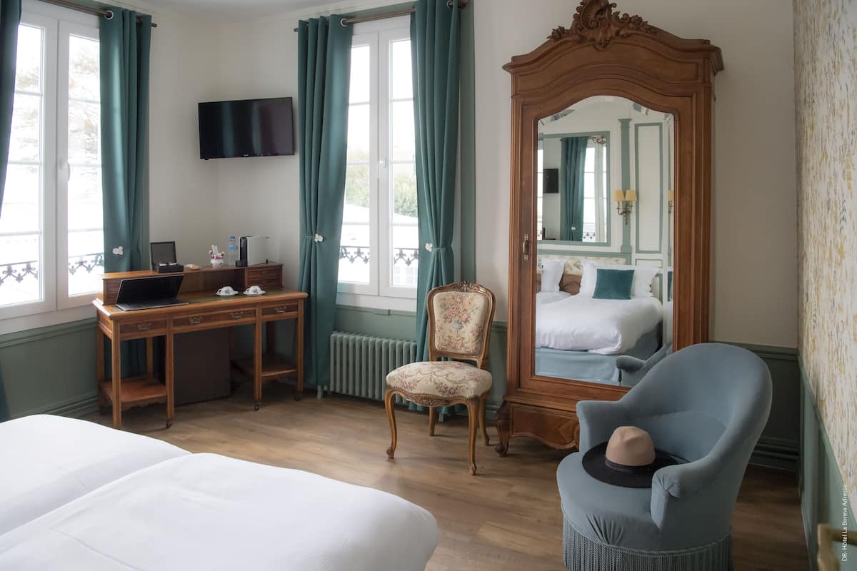 Quarto de hotel com uma secretária de madeira, uma cadeira, uma cama, um espelho grande e uma televisão na parede.