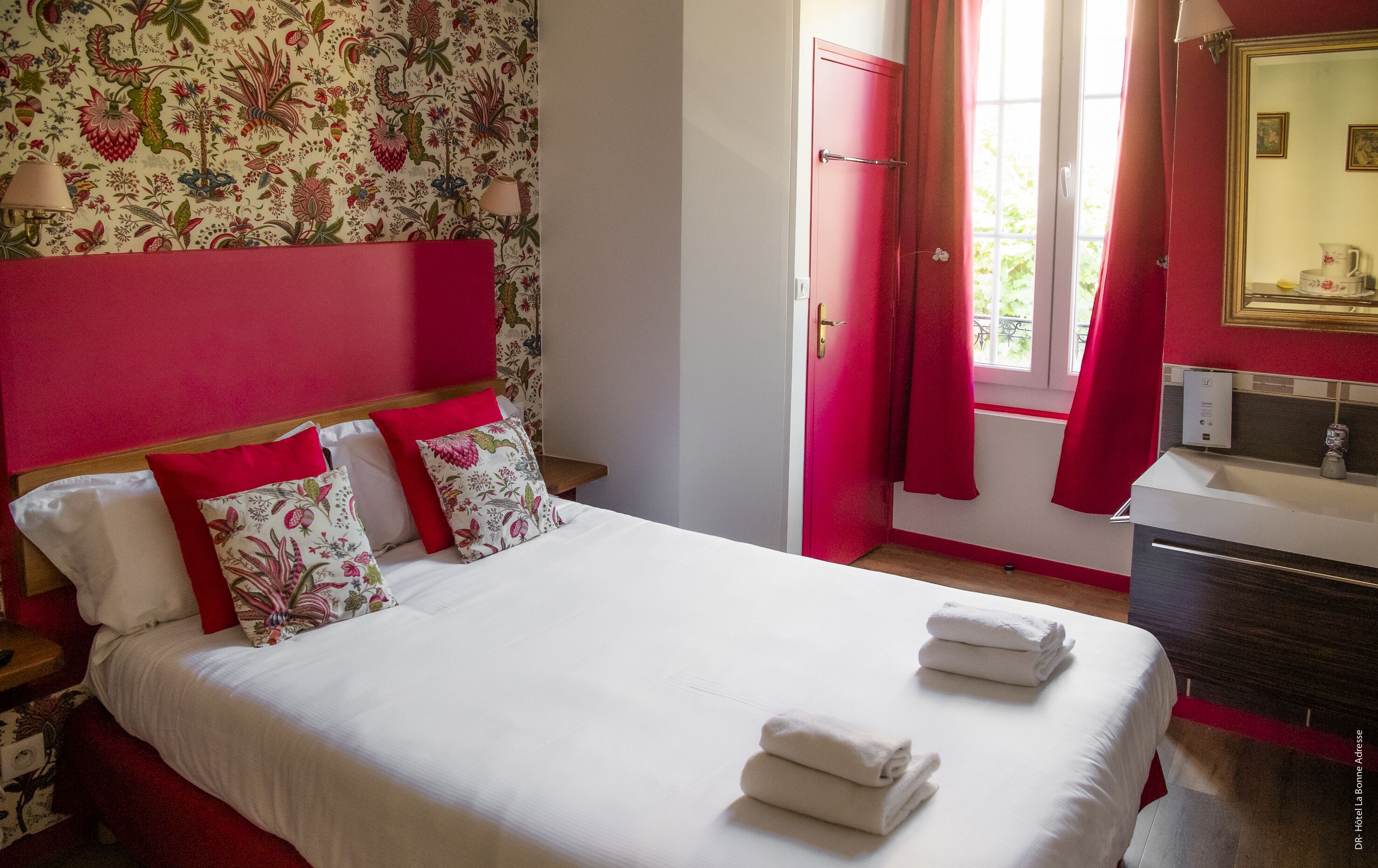 Quarto de hotel com cama, almofadas, mesa de cabeceira, penteadeira, espelho e janela com cortinas.