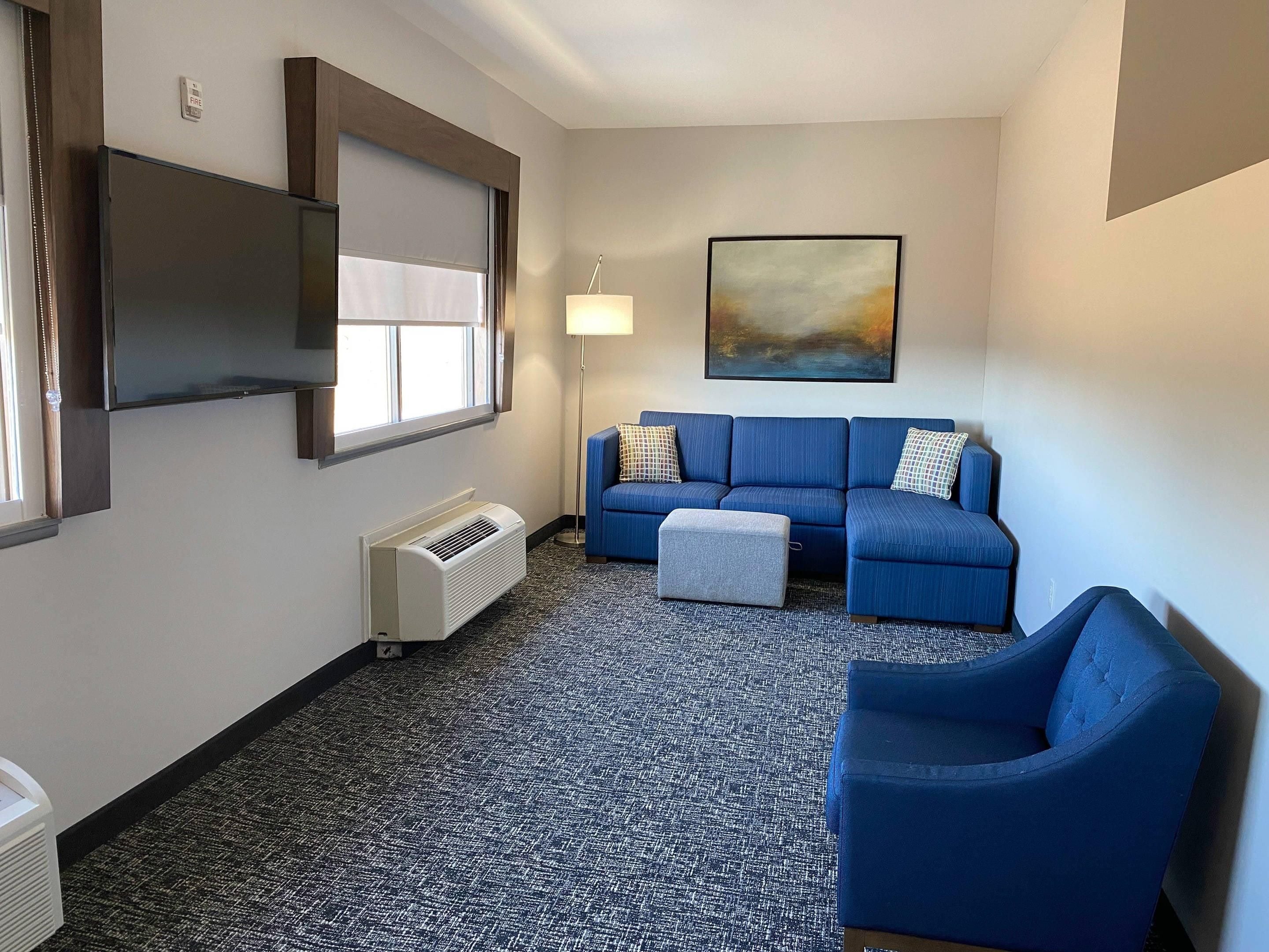 Uma sala de estar moderna com um sofá azul, uma poltrona combinando, um pequeno banquinho, um abajur de chão e um quadro na parede.