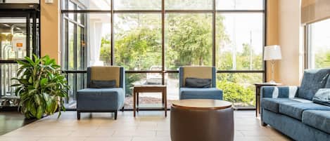 Um lobby moderno com grandes janelas, área de estar com sofás e cadeiras, uma mesa de centro redonda e uma planta em vaso.