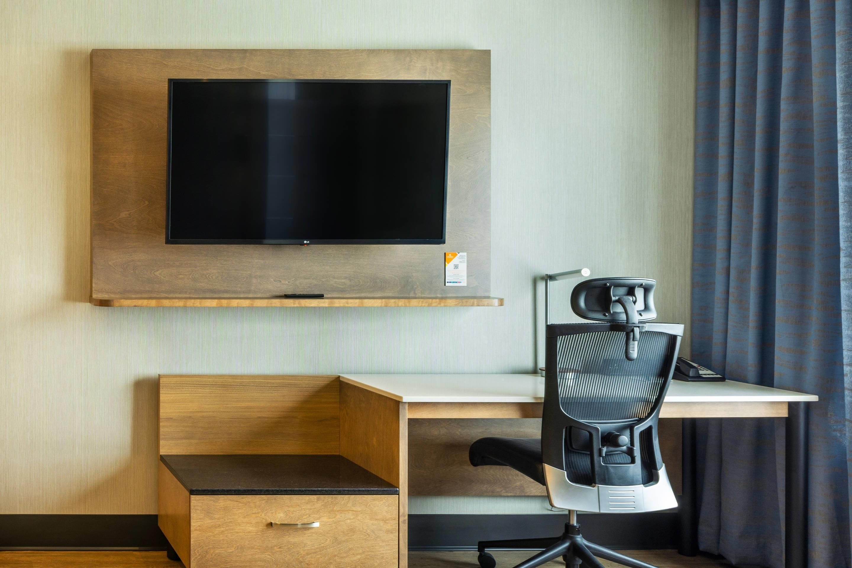 Quarto de hotel com televisão de tela plana na parede, uma escrivaninha de madeira com cadeira e um móvel com gavetas.