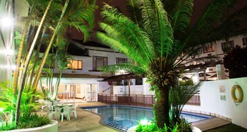 Piscina exterior rodeada por palmeiras e uma área de estar coberta.