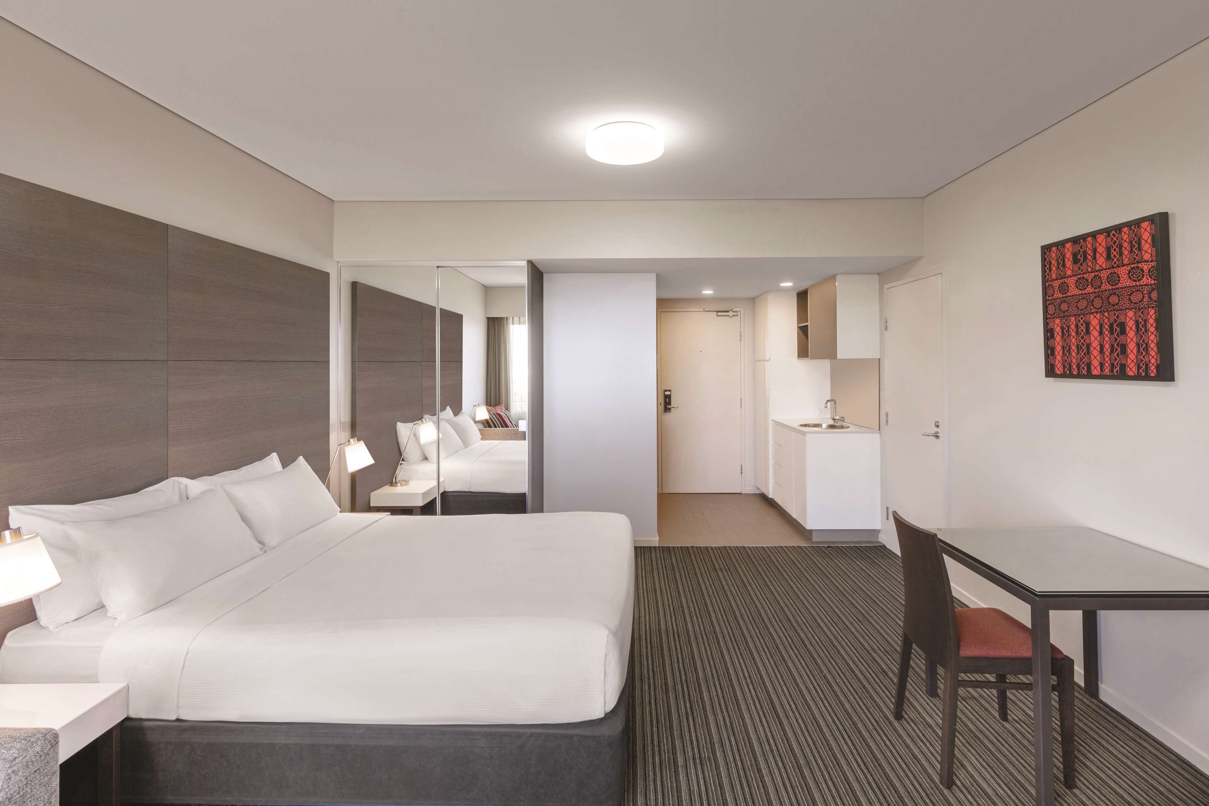 Quarto de hotel com uma cama grande, uma escrivaninha, uma cadeira, uma pequena cozinha e um banheiro visível por uma porta aberta.