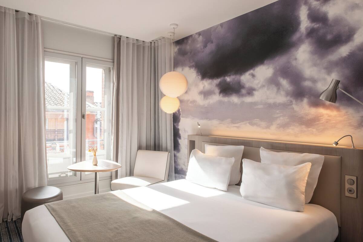Quarto de hotel moderno com uma cama grande, uma mesa redonda pequena, uma cadeira e vista da cidade pela janela.