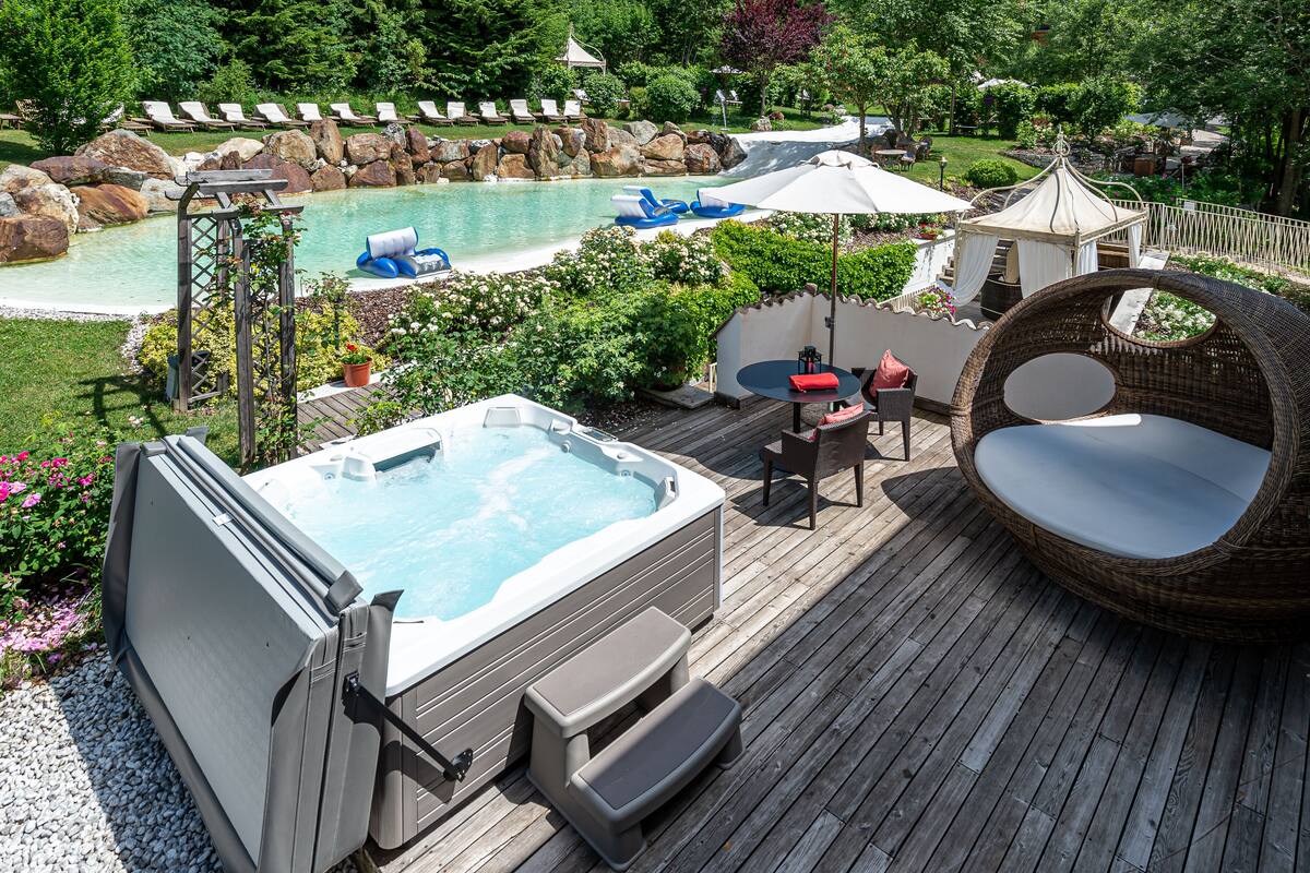 Uma banheira de hidromassagem em um deck de madeira, com uma piscina ao fundo, rodeada por espreguiçadeiras e um gazebo.