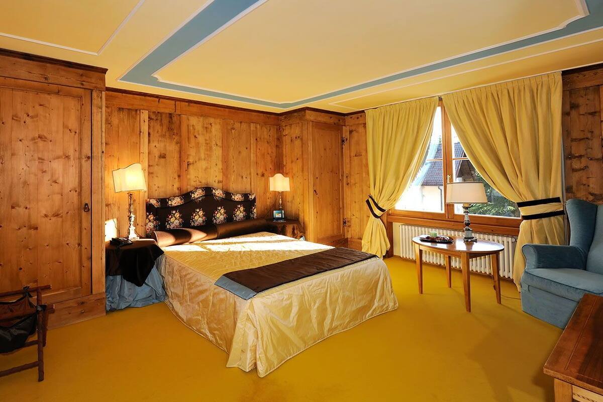 Quarto com painéis de madeira, uma cama, duas mesas de cabeceira, uma cadeira, uma mesinha e uma janela com cortinas.
