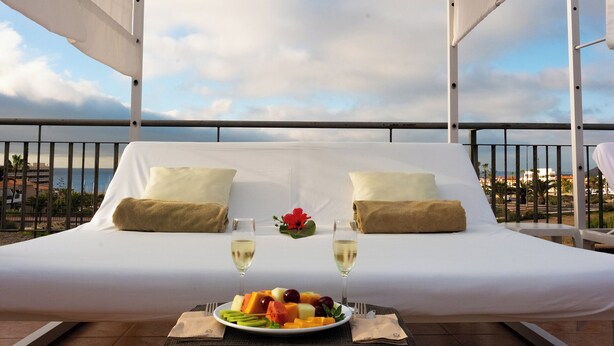Uma cama com duas almofadas, um prato de fruta e dois copos de vinho branco numa varanda com vista.