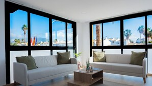 Sala de estar com dois sofás, uma mesa de centro e janelas amplas com vista para palmeiras e edifícios.