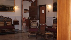 Quarto tradicional com uma grande cadeira de madeira, um sofá e uma pequena mesa. Há um quadro na parede e uma porta que dá para outro cômodo.