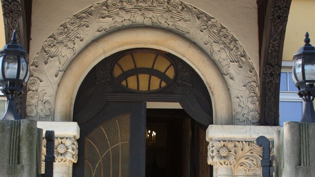 Uma entrada ornamentada com uma grande porta preta, um arco de pedra e um pequeno vaso com planta.