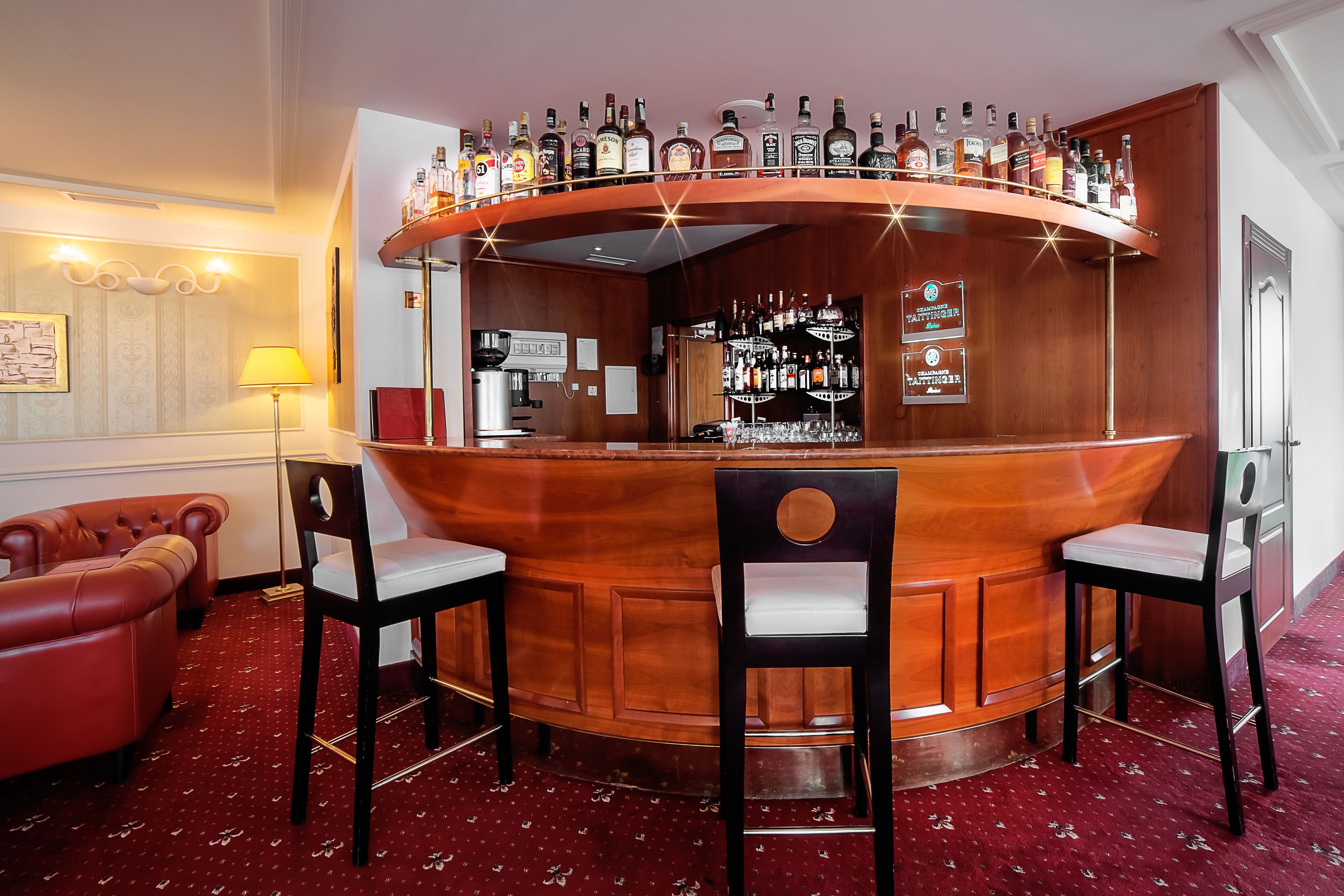 Uma área de bar com balcão de madeira curvo, dois bancos altos e diversos frascos de bebidas alcoólicas.