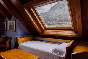Um quarto pequeno e acolhedor com teto inclinado, uma cama de solteiro, uma mesa de madeira e uma janela com vista para uma montanha nevada.