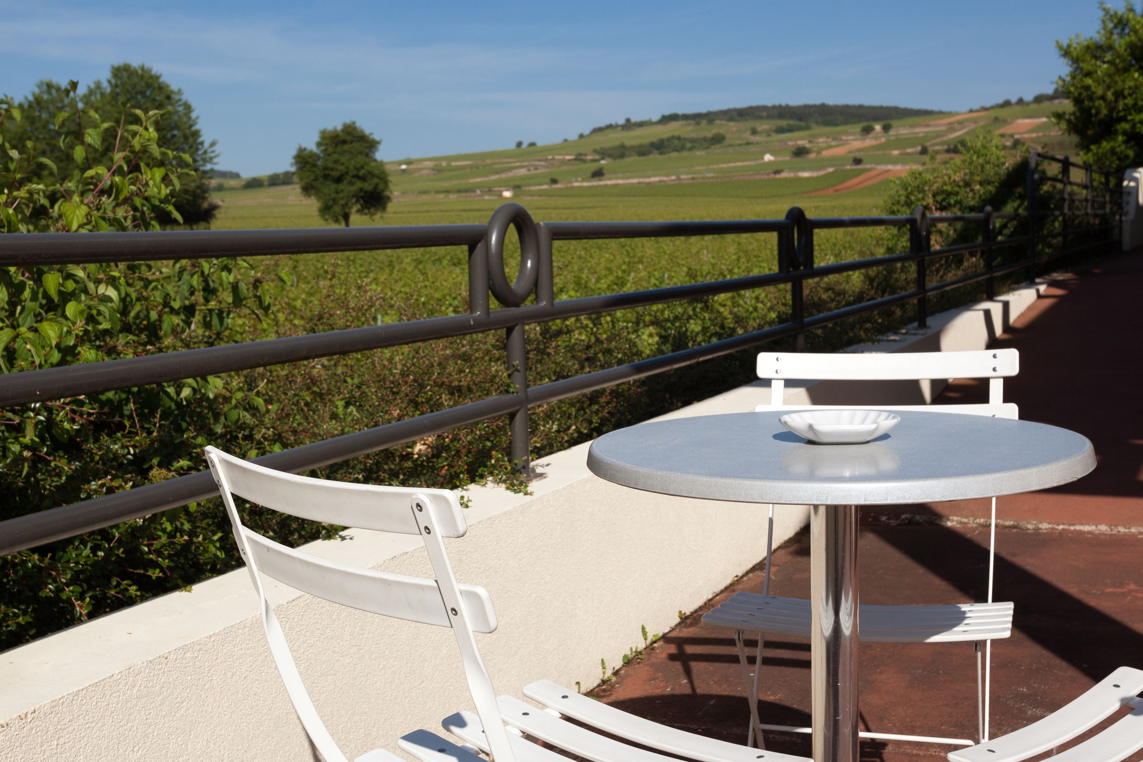Conjunto de cadeira e mesa de metal branco em terraço, com vista para um campo verde e colinas.