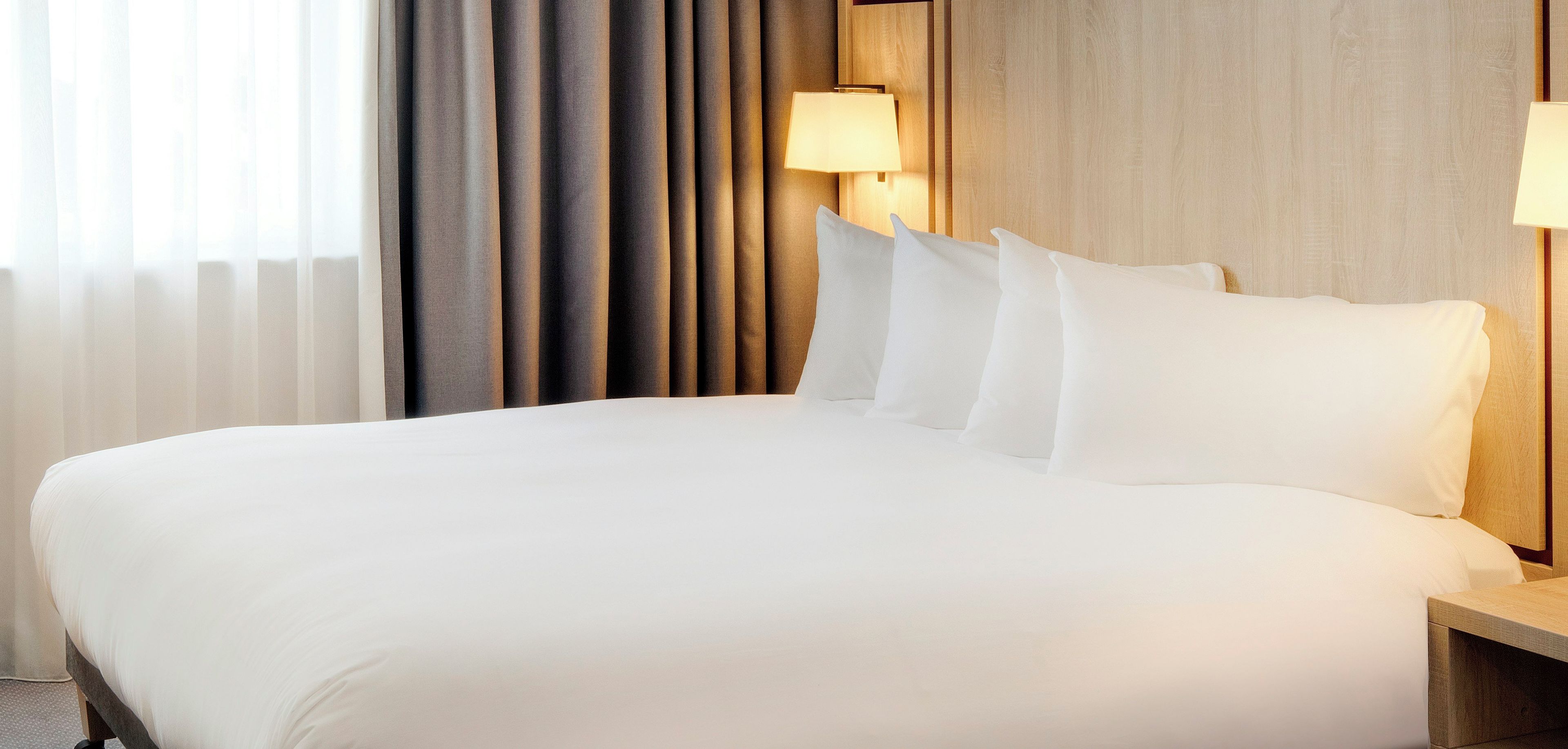 Quarto de hotel com uma cama grande, cabeceira de madeira e duas luminárias de apoio.