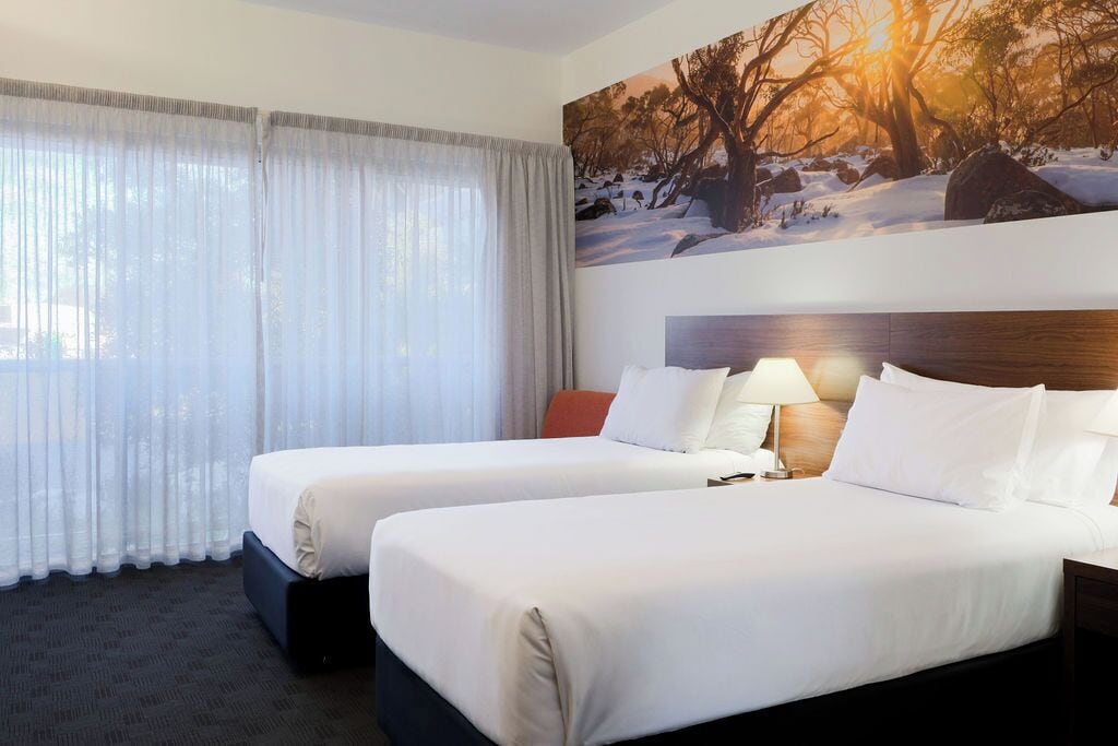 Quarto de hotel com duas camas, uma janela grande com cortinas finas, um quadro de uma paisagem nevada e uma mesa de cabeceira com uma luminária.