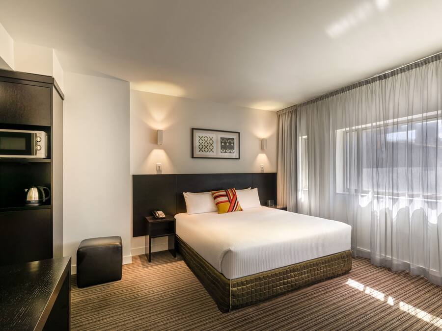 Quarto de hotel com uma cama grande, escrivaninha, micro-ondas, mesa de cabeceira e janela com cortinas finas.