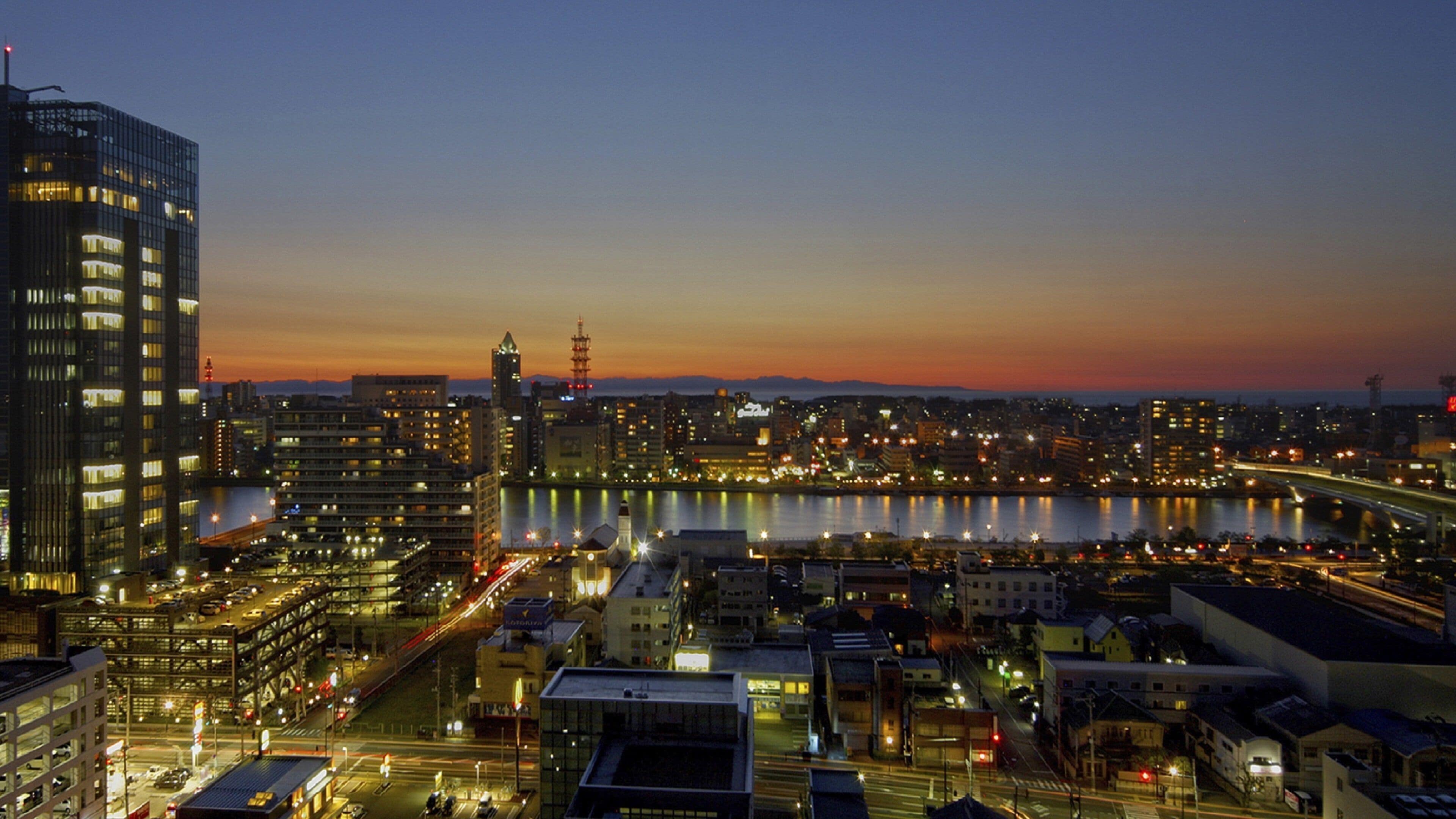 Paisagem urbana ao entardecer, com edifícios iluminados, um rio e uma ponte.