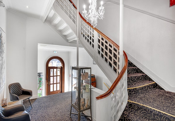 Uma escadaria imponente com corrimão branco e um lustre, que leva a um espaço com carpete estampado e um expositor de vidro.