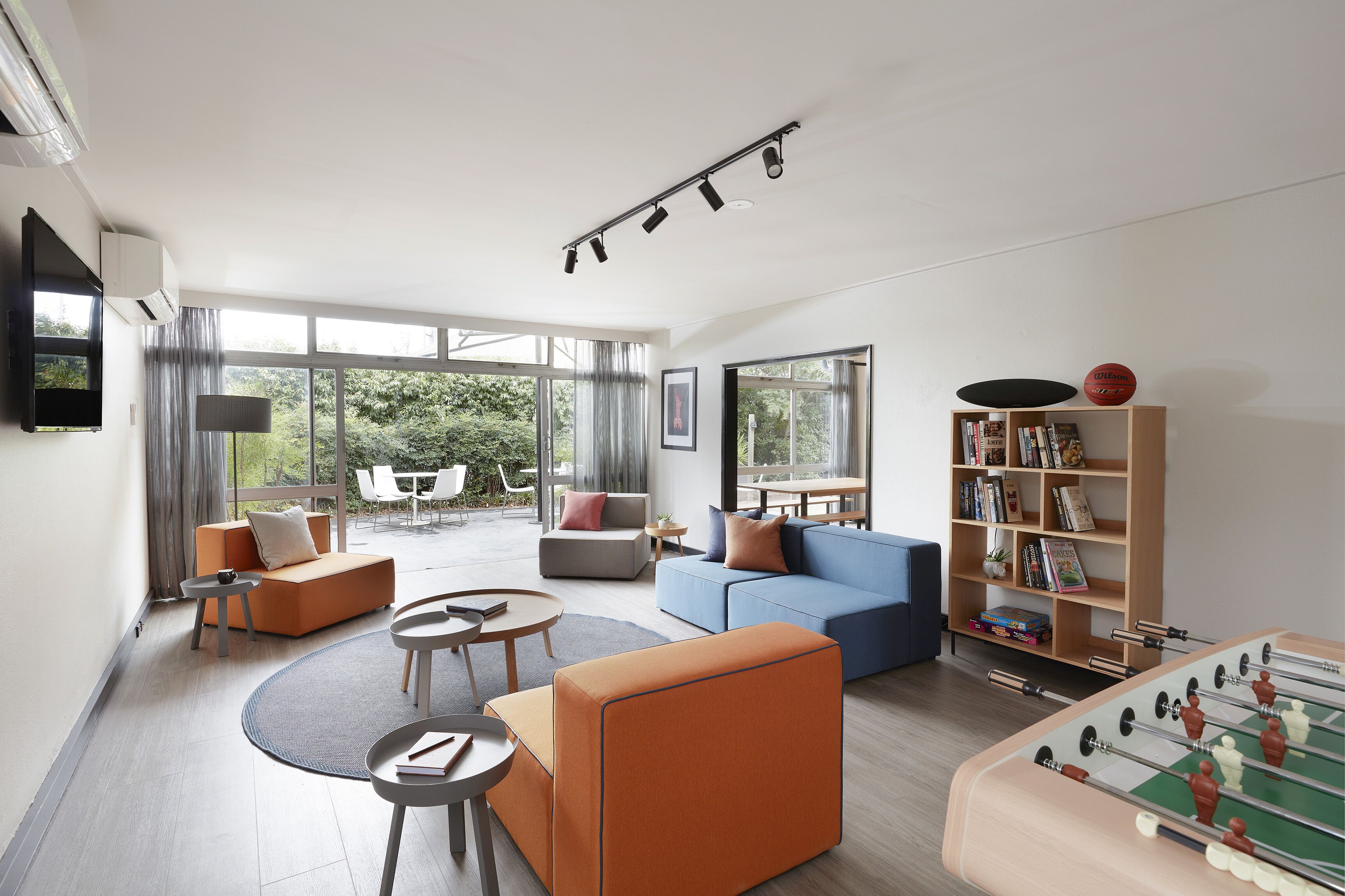Uma sala de estar moderna com um sofá, um banquinho, uma mesa de centro e uma mesa de futebol de mesa. Há uma estante de livros com livros e uma bola de basquete.
