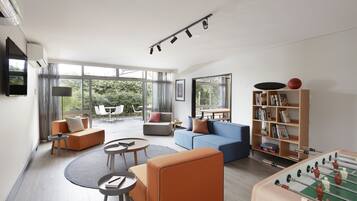 Uma sala de estar moderna com um sofá, um banquinho, uma mesa de centro e uma mesa de futebol de mesa. Há uma estante de livros com livros e uma bola de basquete.