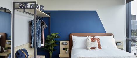 Quarto de hotel moderno com cama beliche, mesa de cabeceira com luminária, planta em vaso e uma grande janela com vista para a cidade.