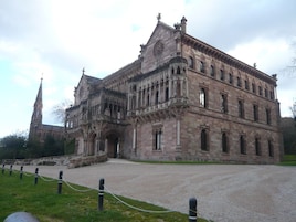 Um edifício histórico com uma grande entrada em arco, uma torre do relógio ao fundo e um pátio pavimentado.