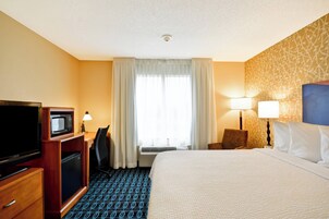 Quarto de hotel com cama, escrivaninha com televisão, micro-ondas, cadeira e luminária.