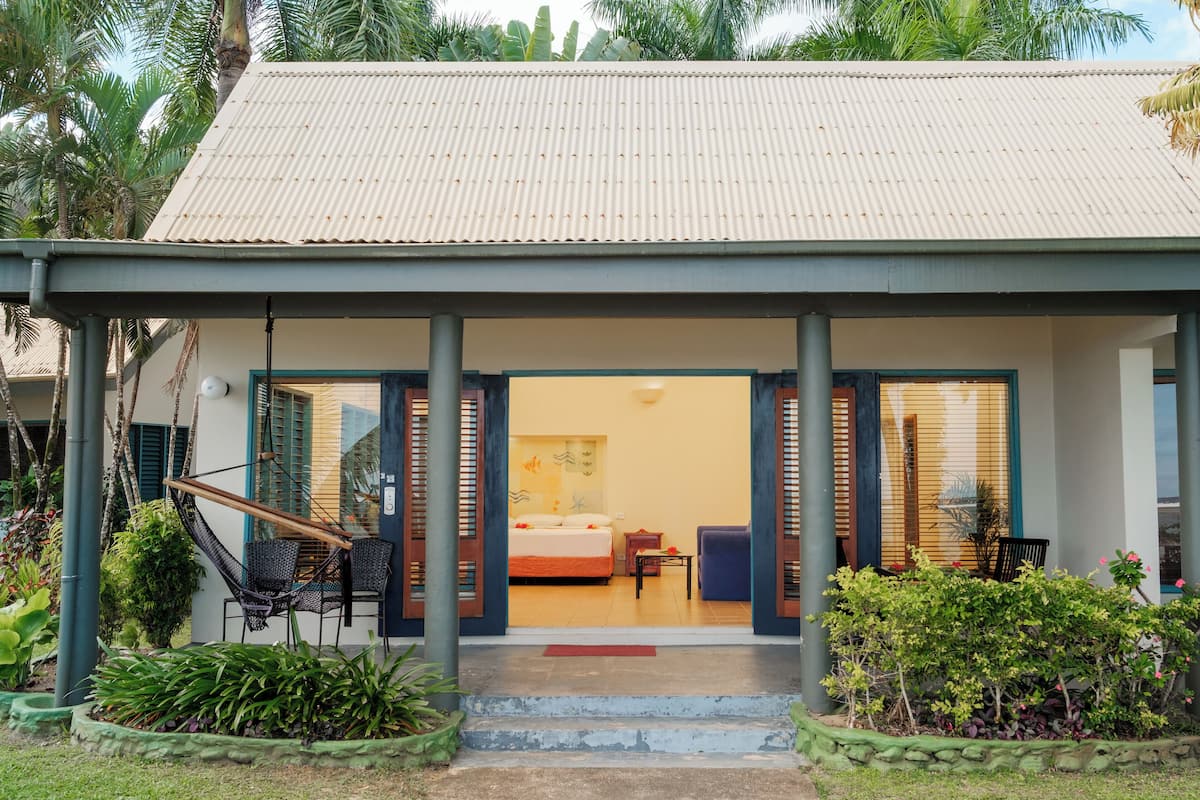 Um hotel com entrada coberta, um quarto com cama e escrivaninha, e um jardim com plantas e uma rede.