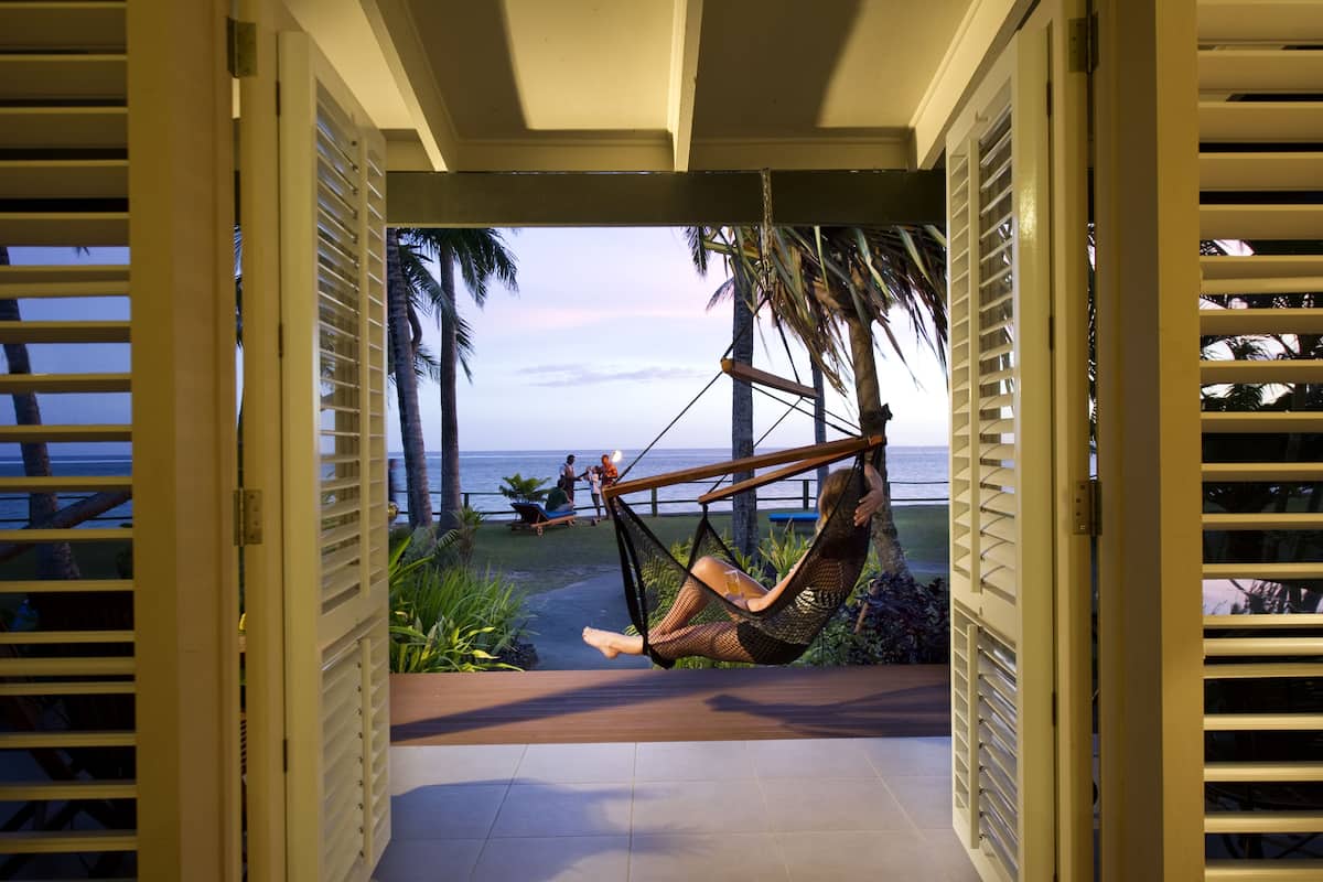 Vista de um quarto com varanda para o oceano, com uma cadeira de rede e um caminho que leva à praia.