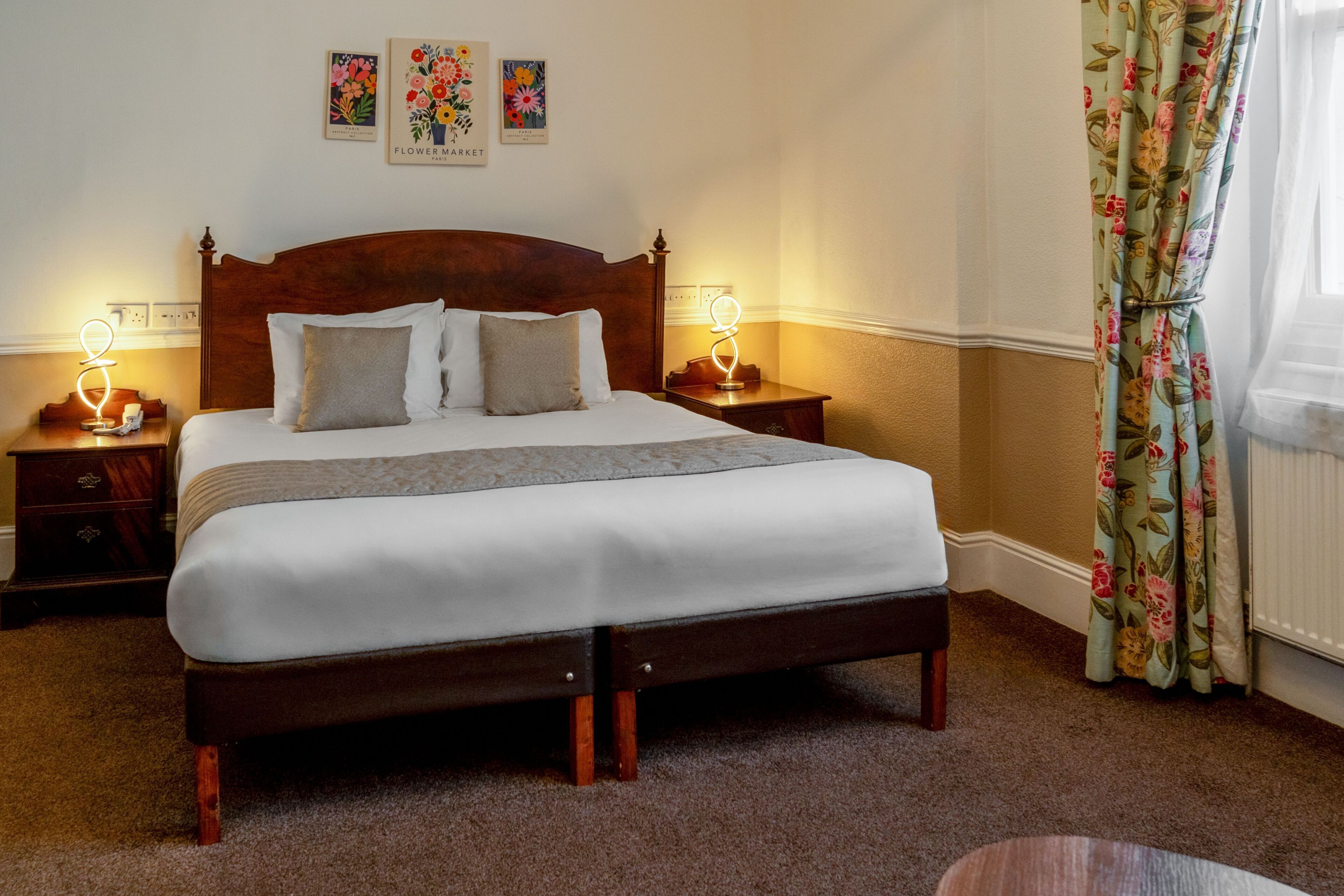 Uma cama bem arrumada com cabeceira de madeira, duas mesas de cabeceira com abajures, e arte floral na parede.