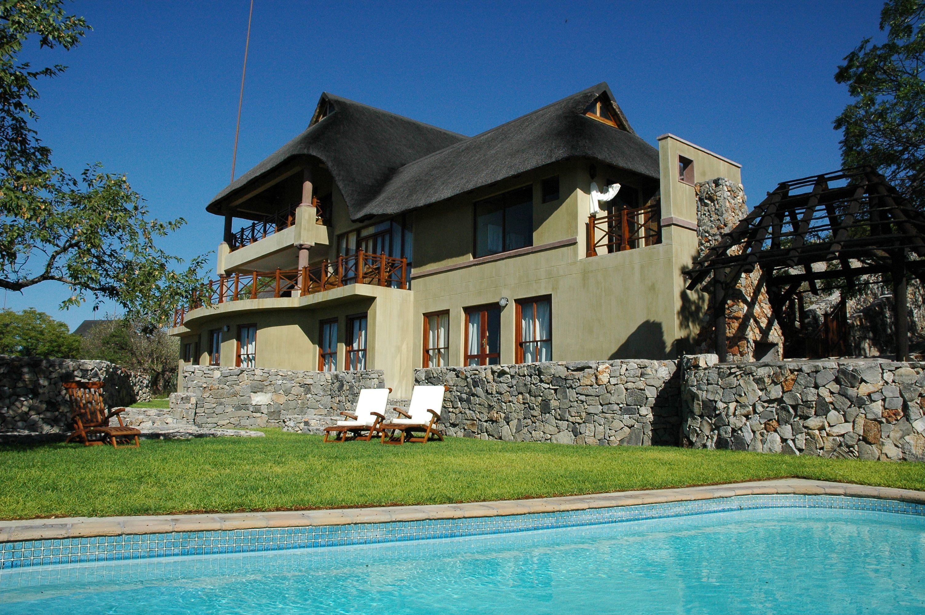 Uma casa de dois andares com fachada de pedra, piscina e telhado de palha.