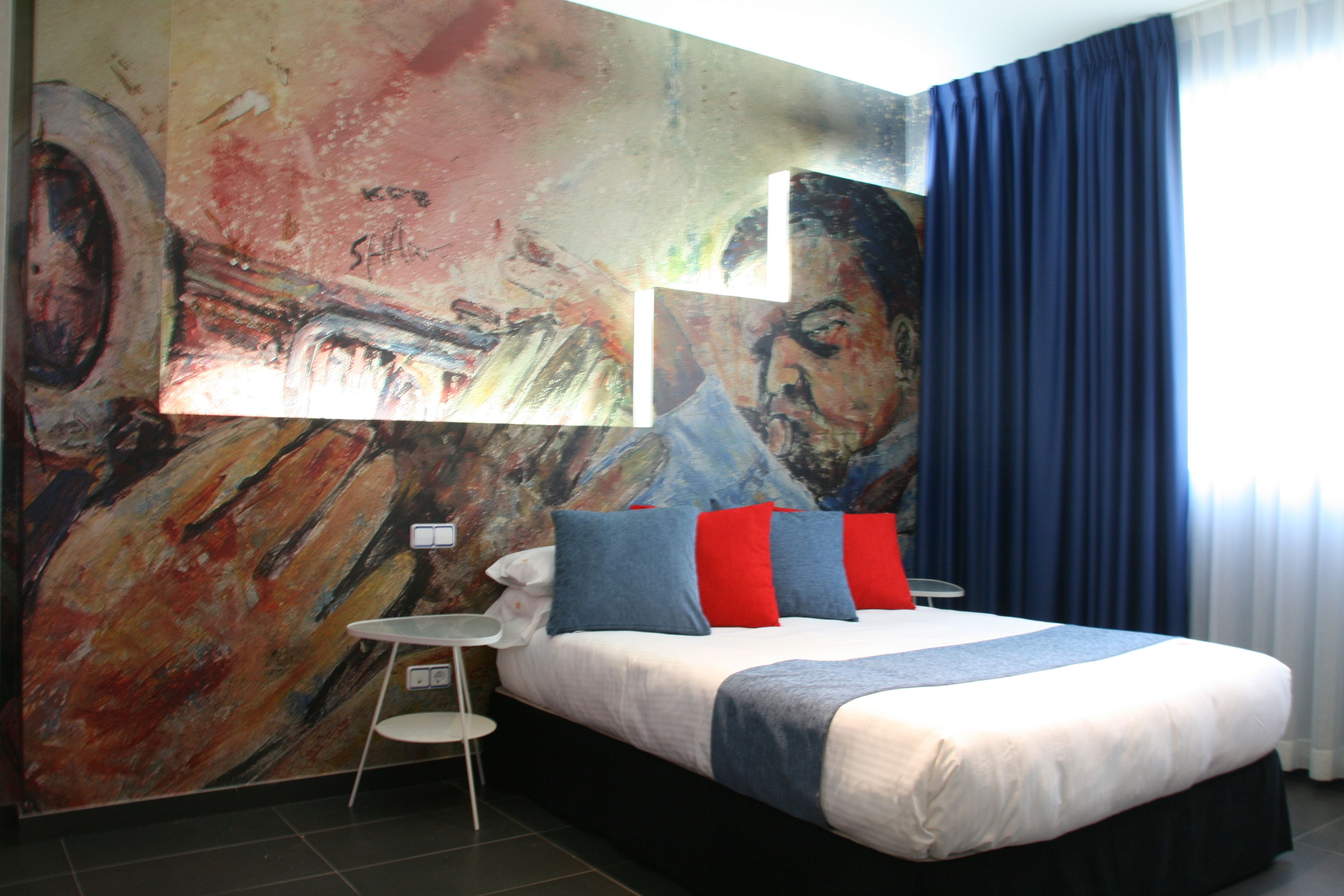 Quarto de hotel moderno com uma grande pintura abstrata na parede, uma cama com almofadas azuis e vermelhas, e uma pequena mesa redonda.