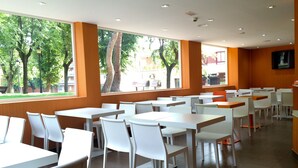 Uma sala de jantar moderna com mesas e cadeiras brancas, grandes janelas com vista para árvores e edifícios, e detalhes em laranja nas paredes.