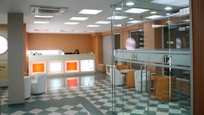 Uma área de recepção com piso xadrez, balcão de atendimento com painéis iluminados e um separador de vidro com assentos.