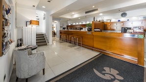 Uma área de recepção de hotel com um balcão de madeira, placa de recepção, relógio e um espaço de bar com banquetas.
