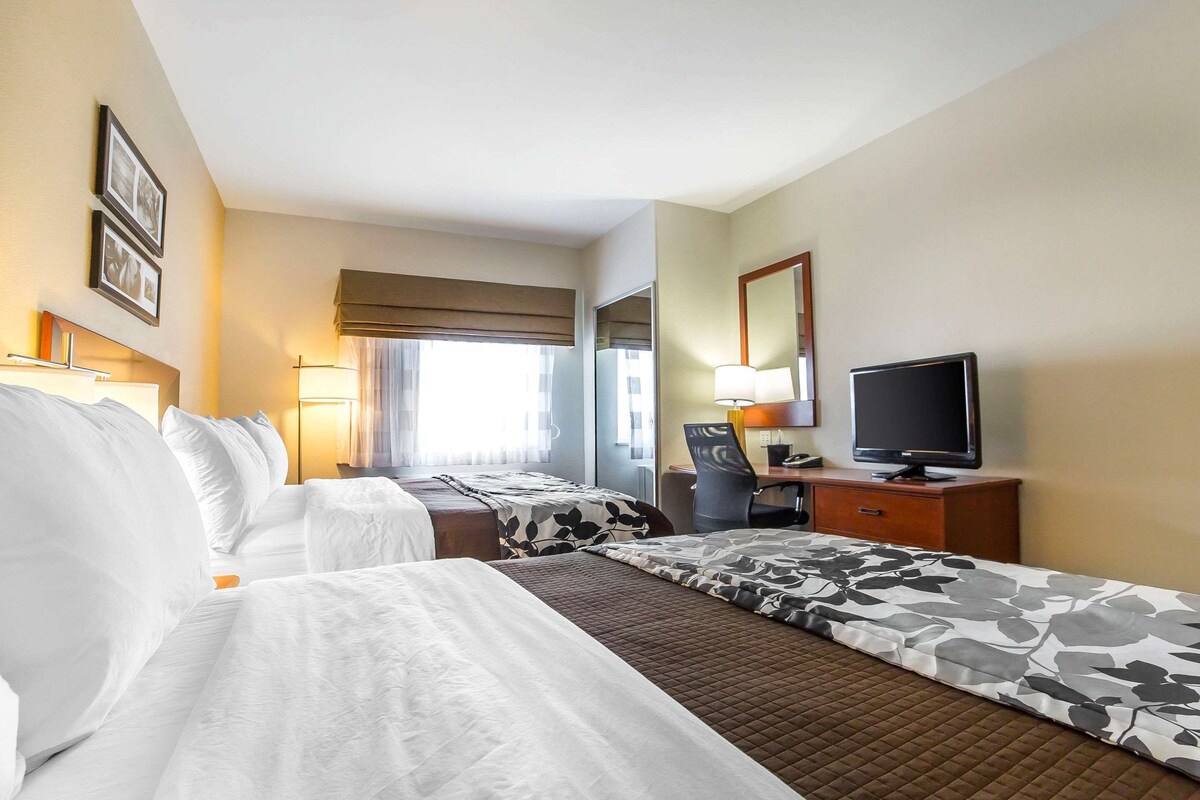 Quarto de hotel com duas camas, uma mesa de trabalho com computador, TV e janela com cortinas.