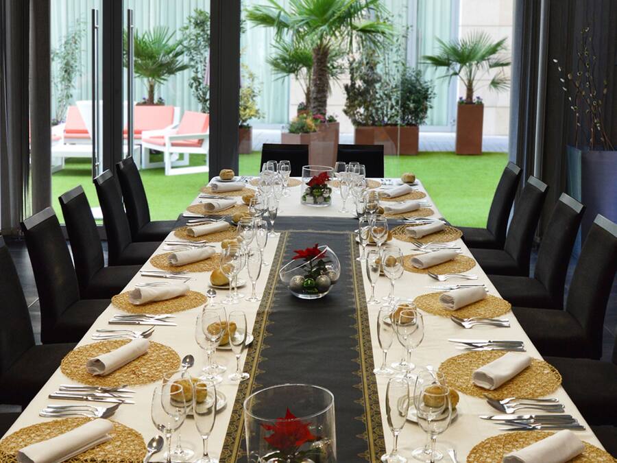 Uma longa mesa de jantar posta para uma refeição formal, com cadeiras em ambos os lados, vista para a área externa com plantas e um edifício ao fundo.