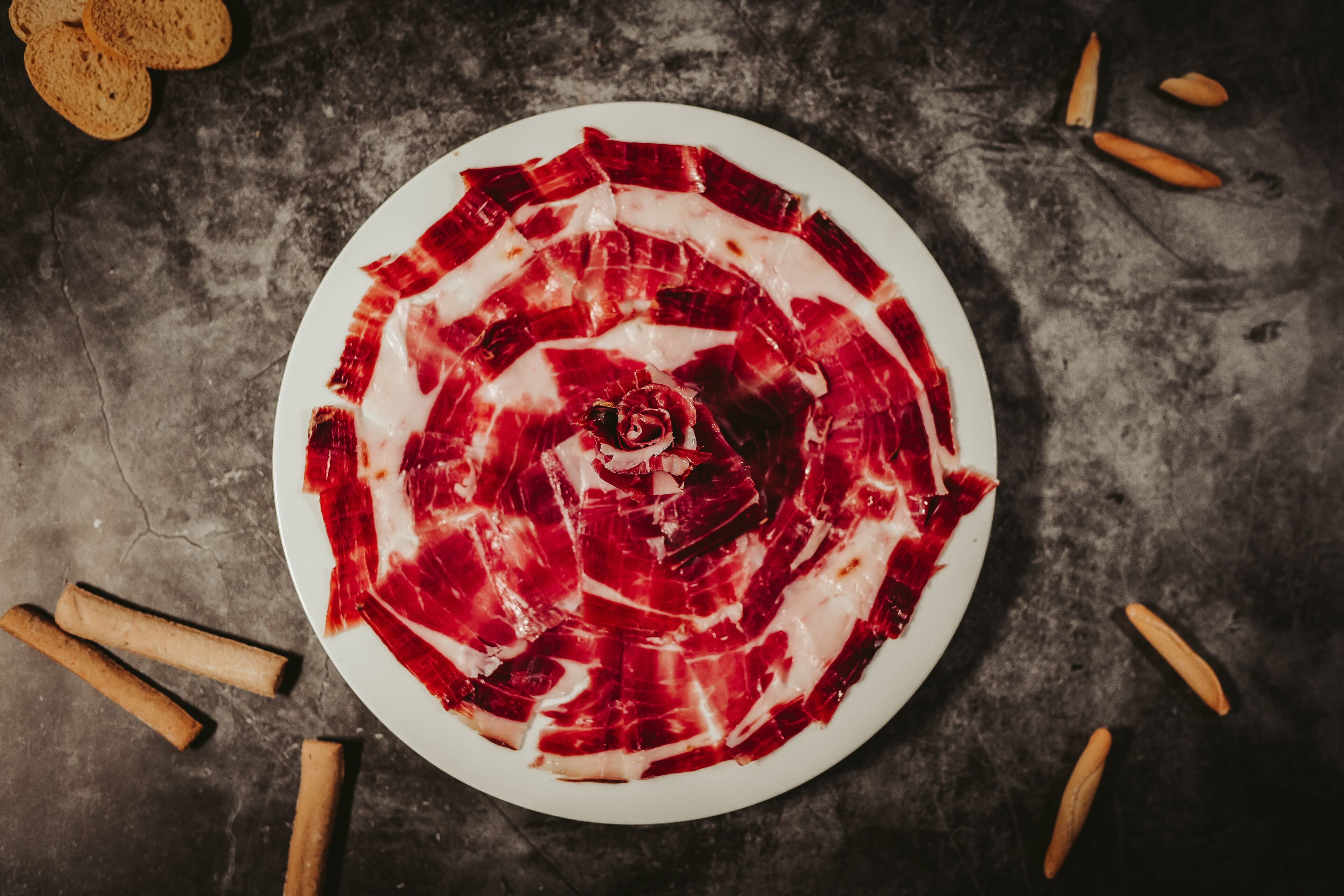 Prato branco redondo com disposição espiral de fatias de carne vermelha e branca, rodeado por palitos de pão espalhados em uma superfície texturizada.