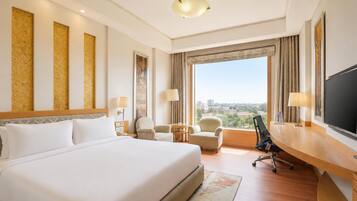 Quarto de hotel com uma cama grande, duas cadeiras, escrivaninha, televisão e janela com vista para a cidade.