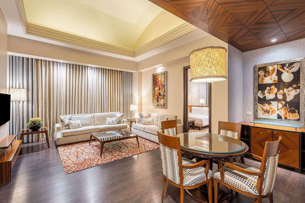 Uma sala de estar moderna com sofá, mesa de centro, mesa de jantar e cadeiras. Há uma janela grande com cortinas, um quadro na parede e um lustre.