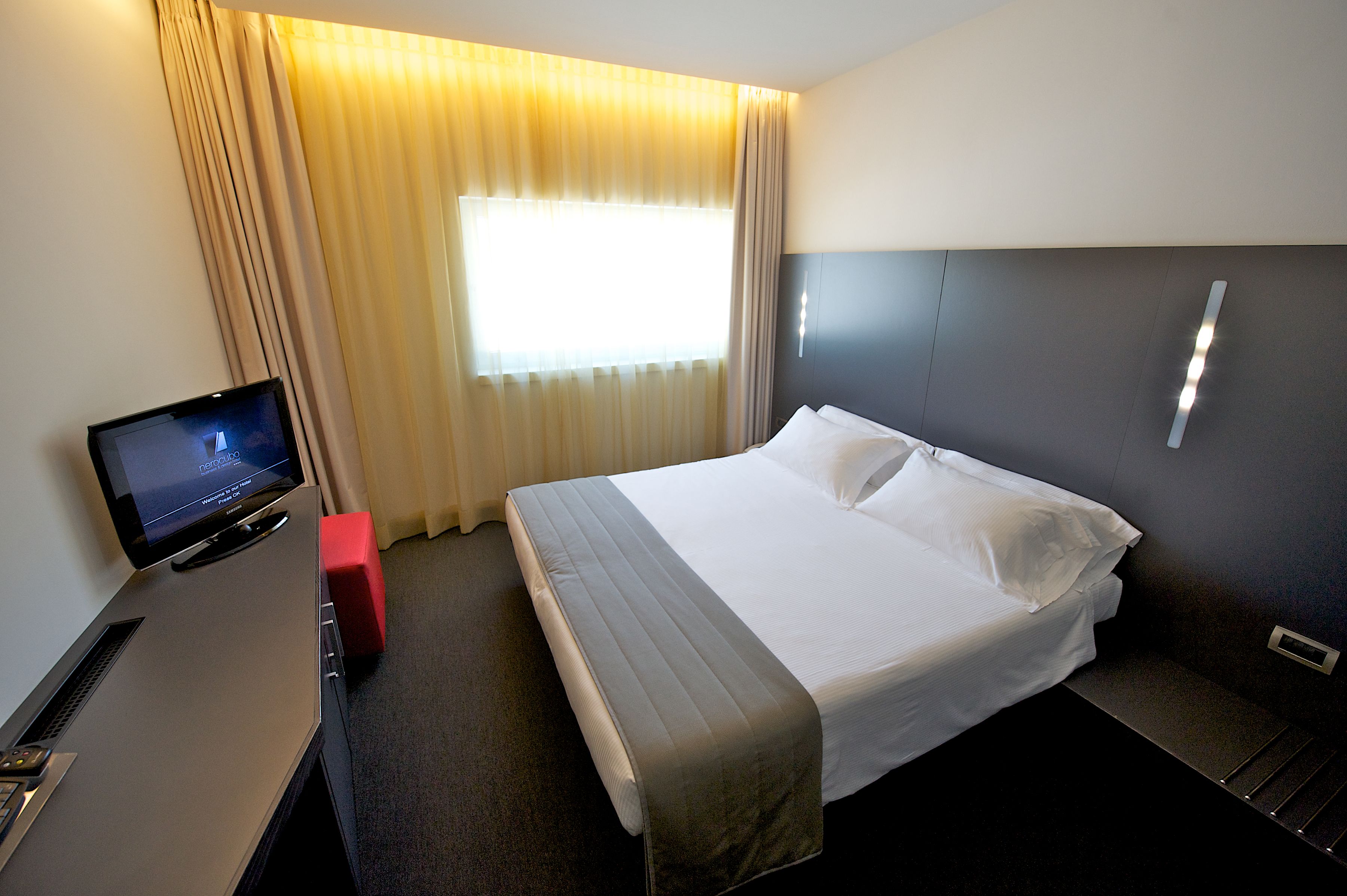 Quarto de hotel com cama, televisão, escrivaninha e janela com cortinas.