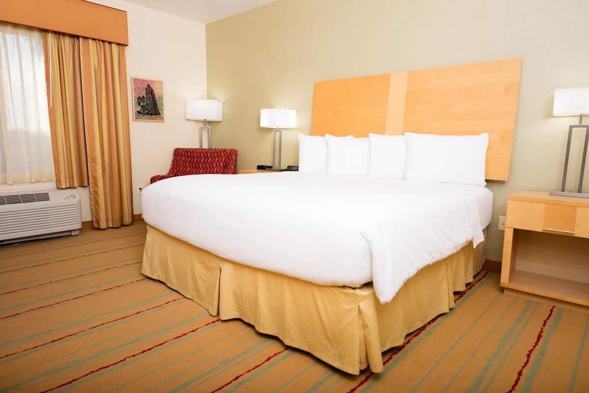 Quarto de hotel com uma cama grande, uma mesa, uma cadeira, um abajur, um quadro na parede e uma janela com cortinas.