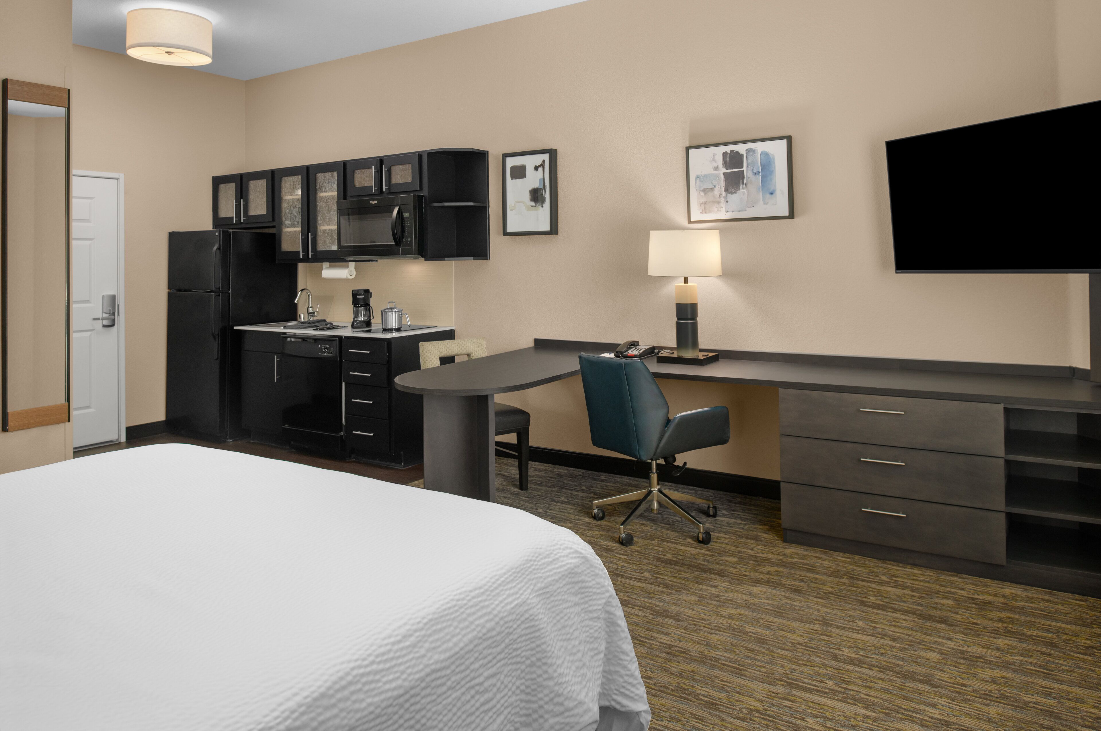 Quarto de hotel com cama, escrivaninha, cadeira, micro-ondas, geladeira, televisão e abajur.