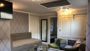 Uma sala de estar moderna com um sofá, poltrona e mesa de centro de vidro. Há uma televisão presa à parede e uma porta que dá para um banheiro.