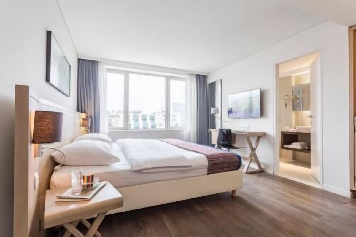 Quarto de hotel com cama, mesa de cabeceira, luminária, TV e vista para a cidade.