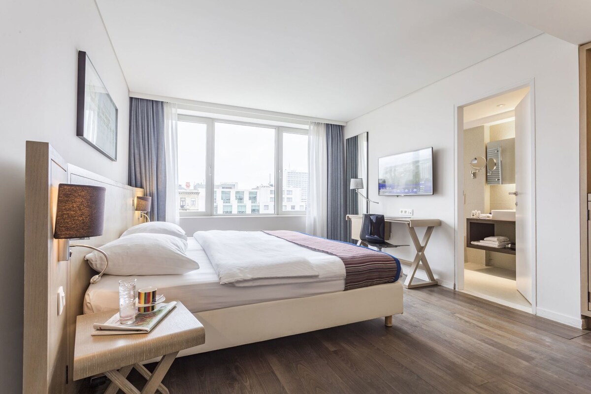 Quarto de hotel moderno com cama, mesa de cabeceira, televisão, escrivaninha e um banheiro visível através de uma porta aberta.