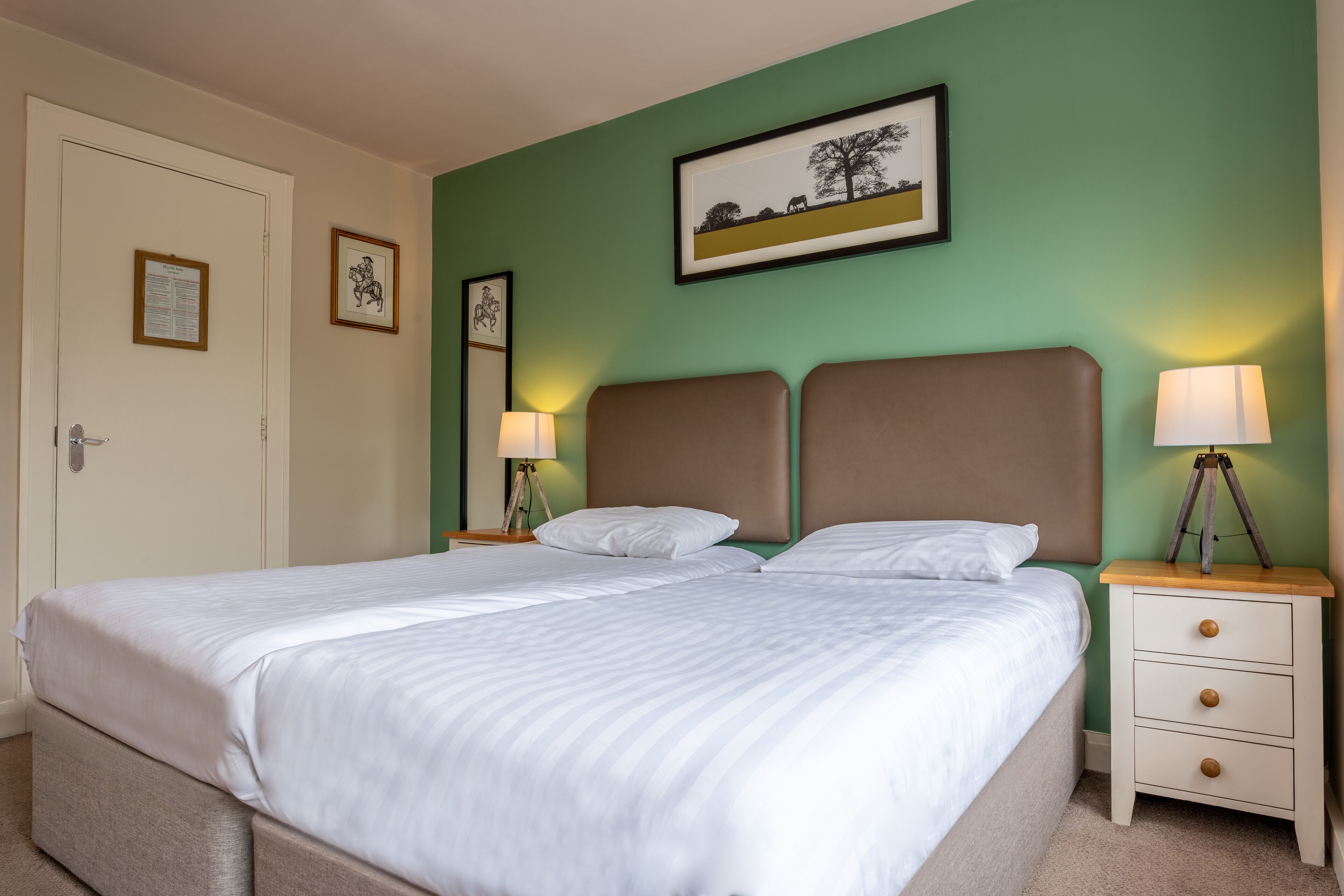 Uma cama de casal com lençóis brancos, uma mesa de cabeceira de madeira com abajur, e uma parede verde com obras de arte emolduradas.